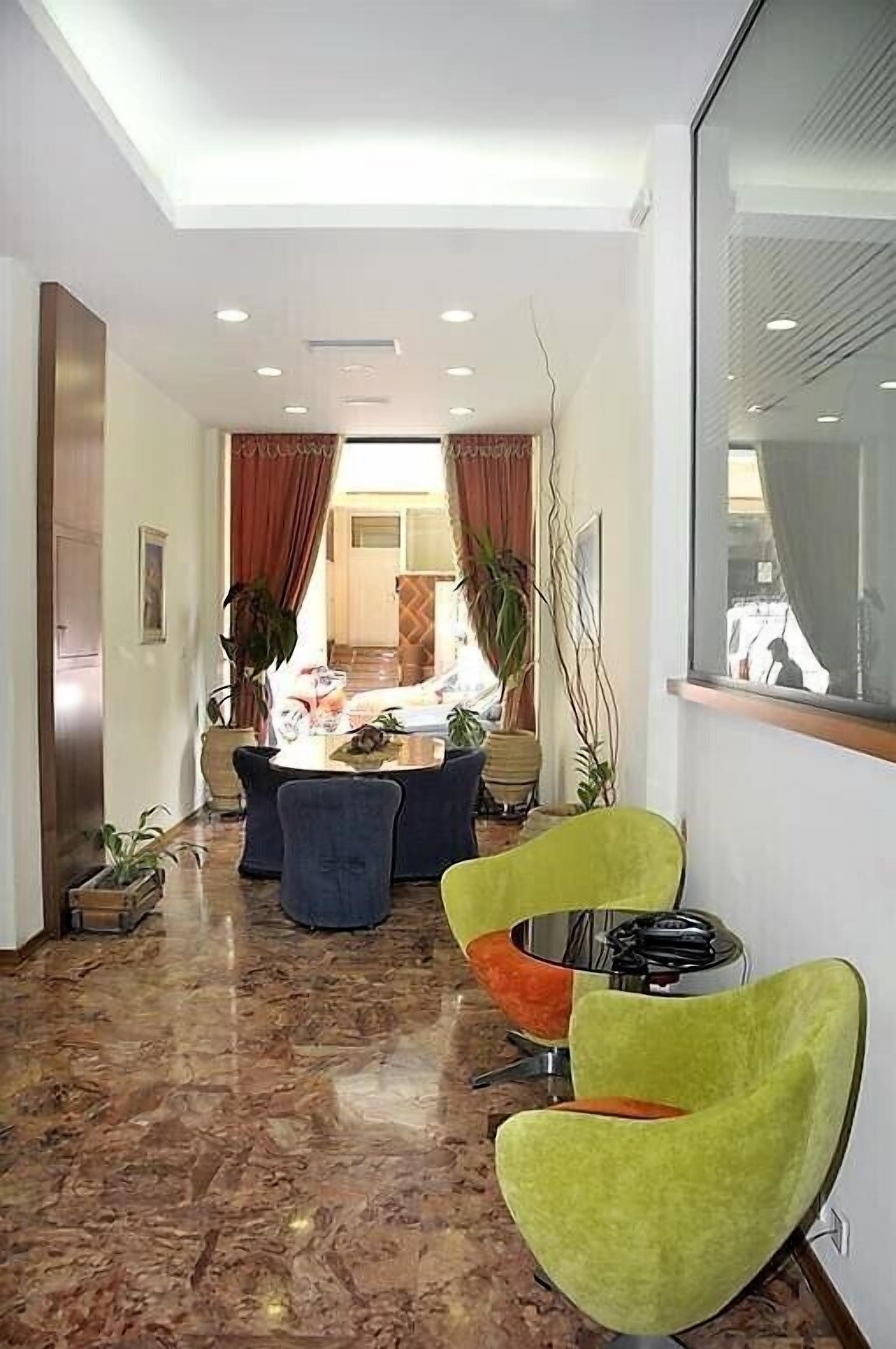 Um hall de hotel moderno com piso de mármore, uma área de refeições com uma mesa redonda e cadeiras, e uma grande janela com cortinas.