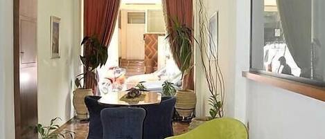 Um hall de hotel moderno com piso de mármore, uma área de refeições com uma mesa redonda e cadeiras, e uma grande janela com cortinas.