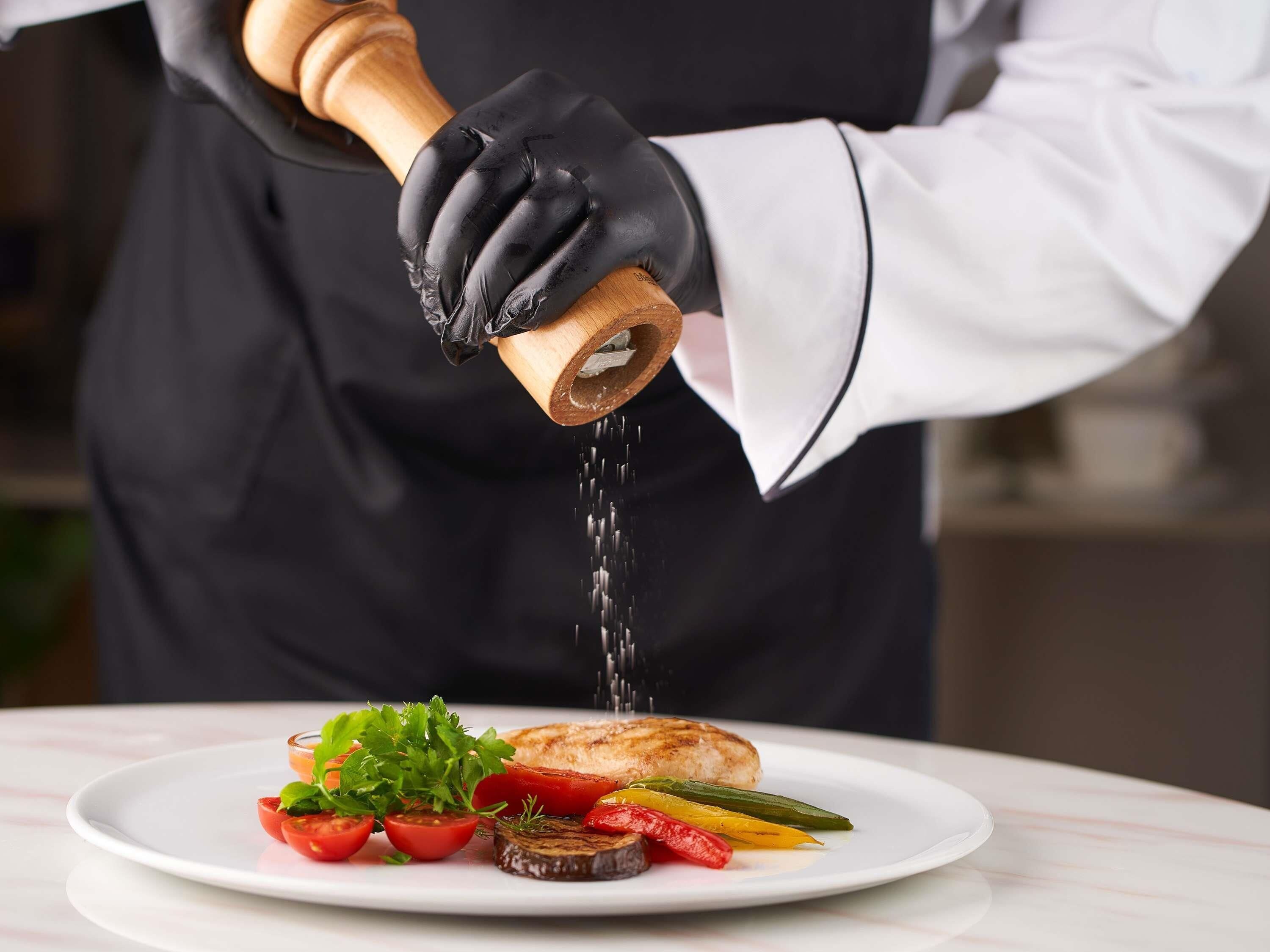 Uma pessoa com avental e luvas pretas despejando água de um jarro de madeira sobre um prato de comida.