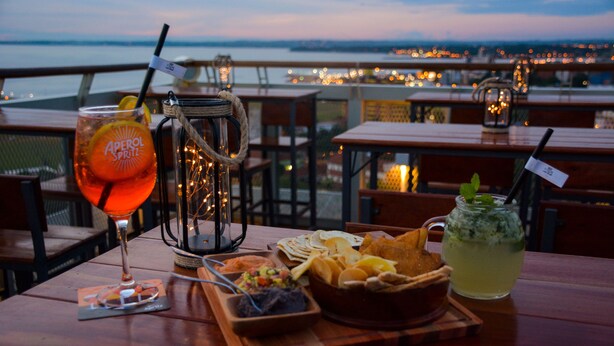 Uma mesa com bebida, jarra e tigela de petiscos em terraço com vista para a água.