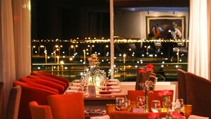 Um restaurante com cadeiras estofadas vermelhas, uma mesa posta com copos de vinho e pratos, e vista para um horizonte urbano iluminado através de uma janela.