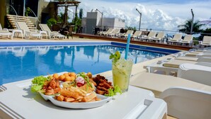 Uma área de refeições à beira da piscina, com uma mesa posta para refeição, incluindo um prato de comida e uma bebida, e cadeiras de descanso ao redor da piscina.