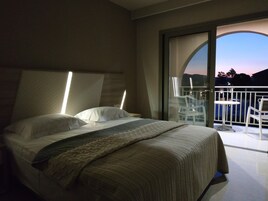 Quarto de hotel com cama, uma janela grande e varanda com cadeiras.