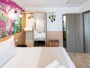 Quarto de hotel com cama, guarda-roupa, um banquinho de madeira, planta pendente e parede com mural tropical.