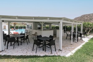 Um pátio externo com mesas e cadeiras, piscina e área coberta com bar.