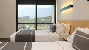 Quarto de hotel com duas camas, uma janela grande com vista para um horizonte urbano e montanhas, e uma luminária moderna fixada na parede.
