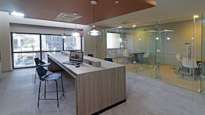 Um espaço de escritório moderno com uma mesa de madeira central, dois bancos altos e uma divisória de parede de vidro.