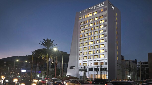 Um edifício alto de hotel com vários andares e janelas iluminadas, cercado por uma rua movimentada com carros e palmeiras.