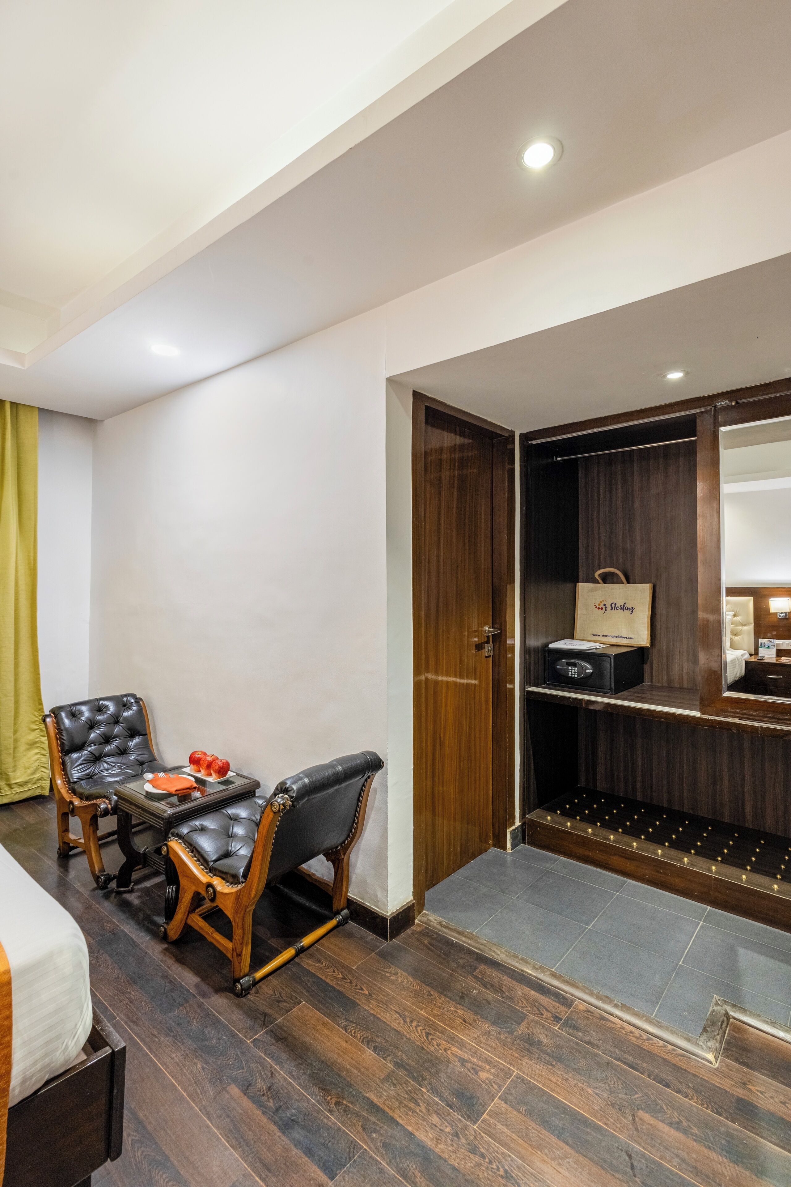Quarto de hotel com piso de madeira, uma mesa de jantar com cadeiras, cama, guarda-roupa e espelho.