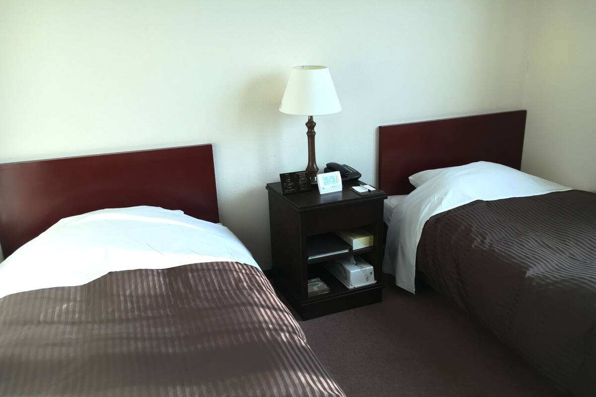 Quarto de hotel com duas camas, uma mesa de cabeceira com abajur, relógio e telefone.