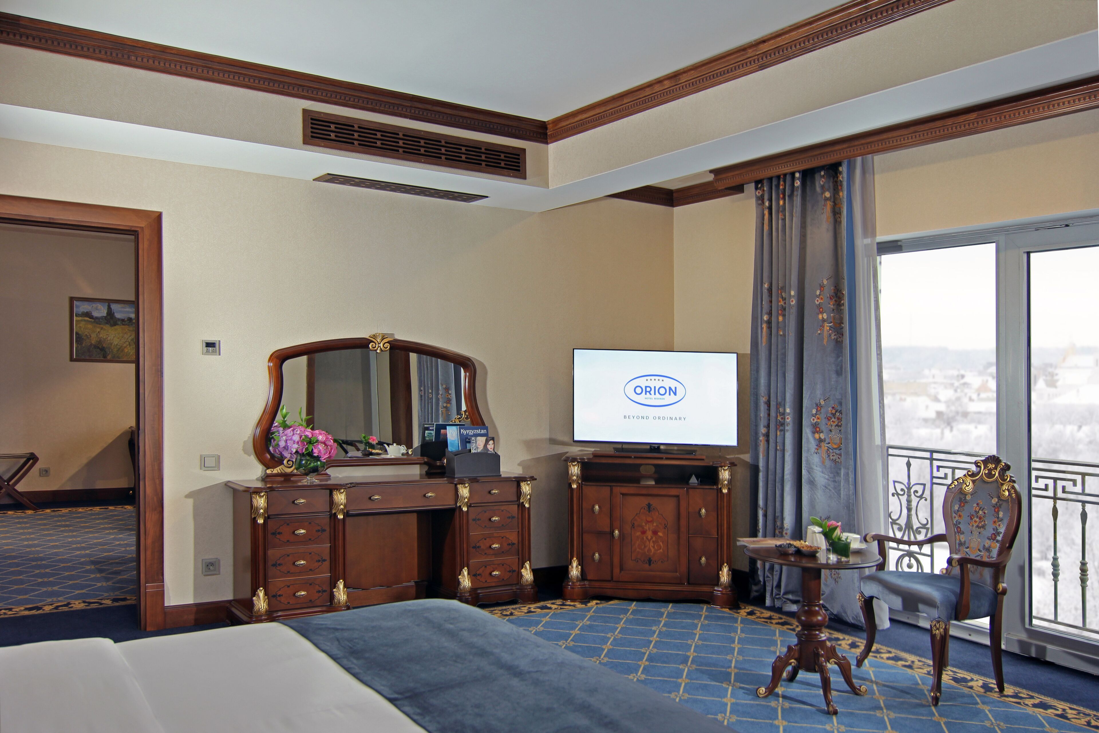 Quarto de hotel com uma cama, cômoda com espelho, televisão, mesa pequena com um vaso e uma cadeira.
