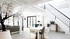 Uma área de refeições moderna com uma mesa e cadeiras brancas, uma grande árvore em vaso e uma escada com corrimão preto.