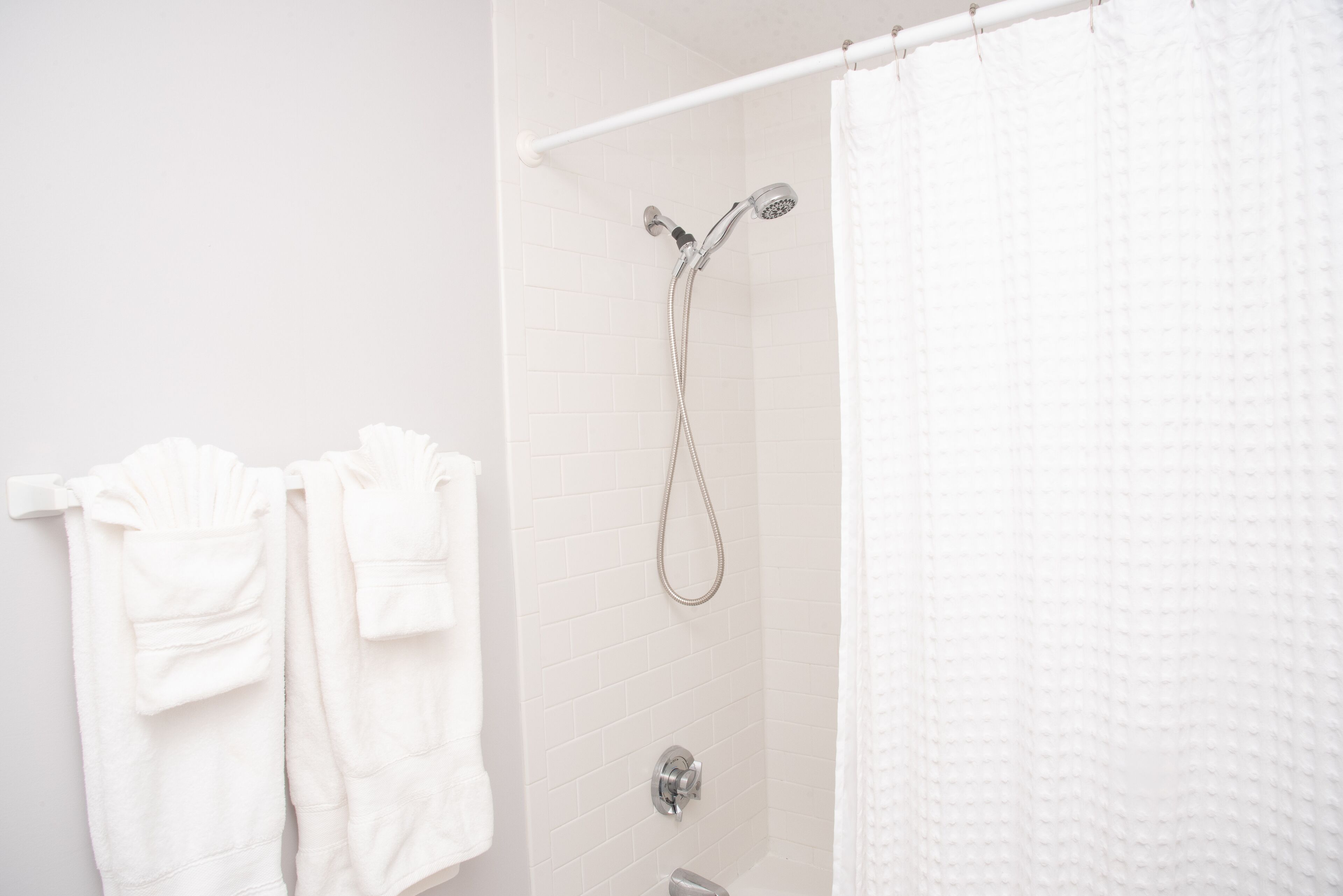 Um chuveiro com chuveiro manual e torneira embutida na parede, um porta-toalhas branco com toalhas, e um box de chuveiro com cortina branca.
