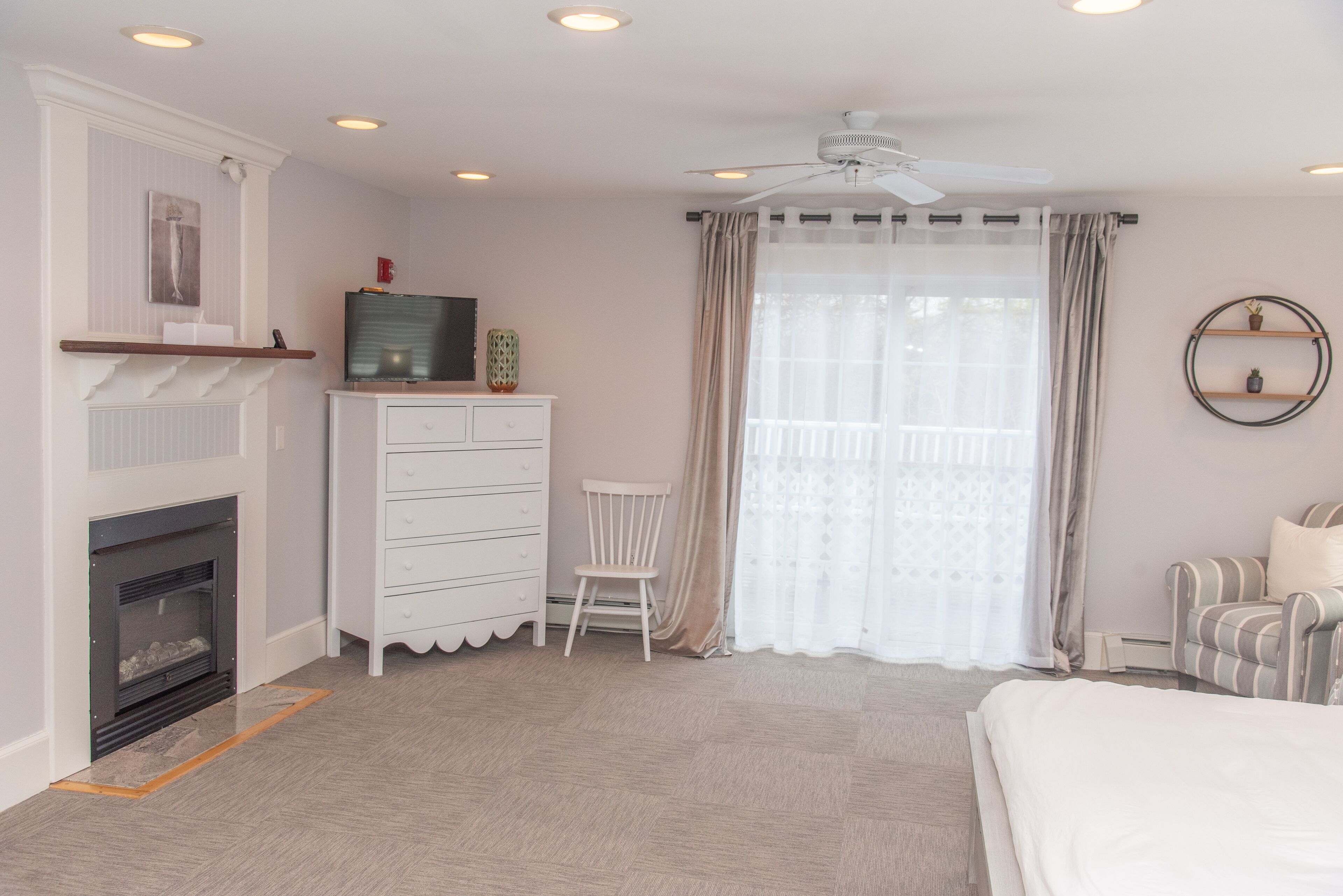Quarto com cama, penteadeira branca, televisão, lareira, ventoinha no teto e janela com cortinas.