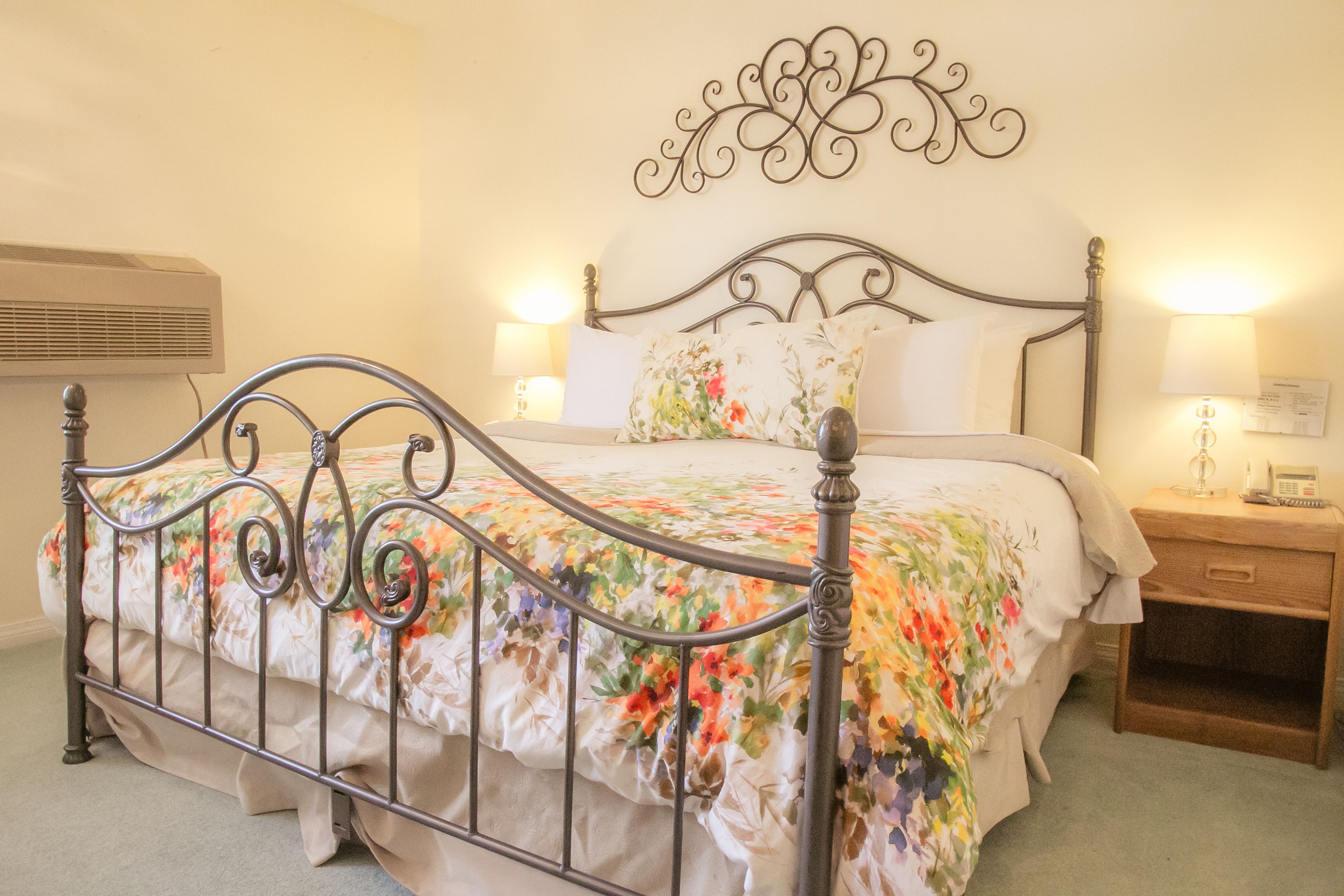 Cama bem arrumada com colcha floral, um objeto decorativo na parede acima e uma mesa de cabeceira com abajur e telefone.