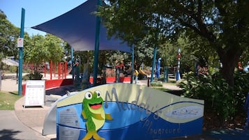 Entrada de um parquinho com um mascote de sapo em desenho animado, área de recreação coberta e árvores ao redor.