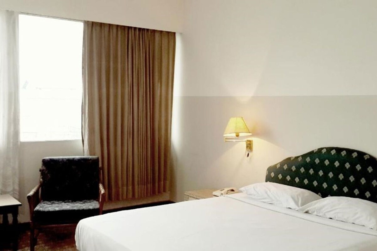 Quarto de hotel com uma cama, uma cadeira, um abajur e uma janela com cortinas.