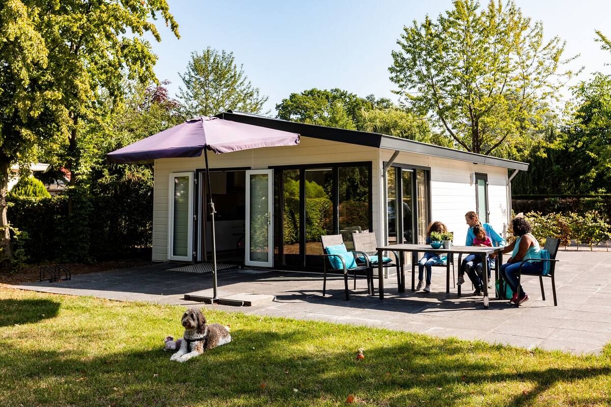 Uma área de varanda coberta com mesa e cadeiras, um cão deitado na grama e um pequeno edifício com portas de vidro.