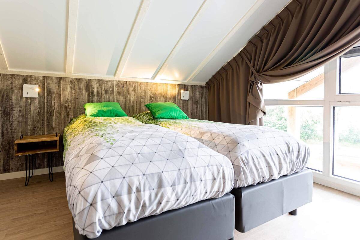 Um quarto com parede de madeira, uma cama com colcha de padrão geométrico, dois travesseiros verdes, uma mesa de cabeceira e uma janela com cortinas.