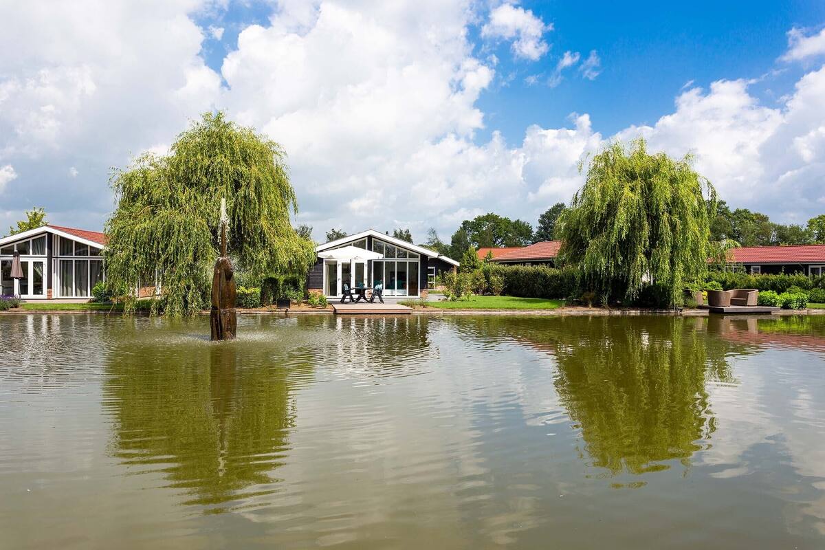 Uma vista tranquila do lago com dois edifícios modernos, um com varanda coberta e outro com área de piscina aparente.
