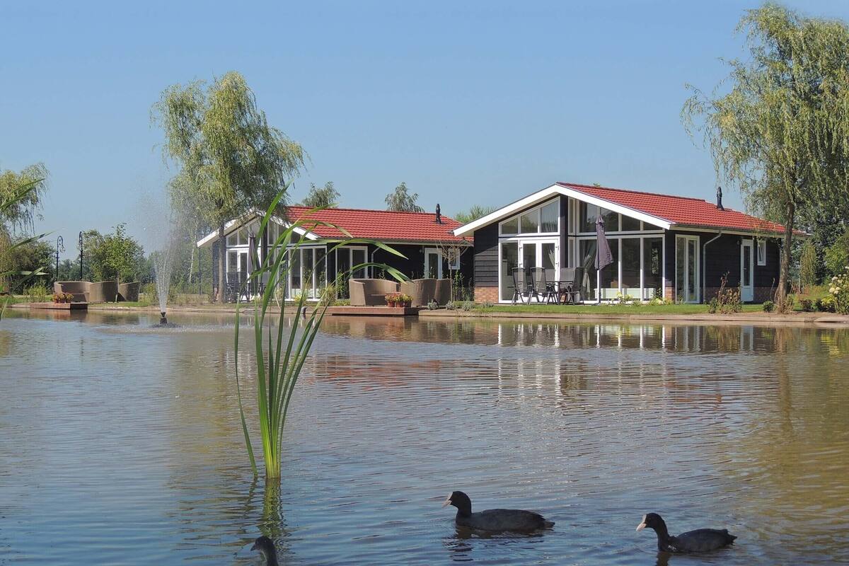Uma casa com telhado vermelho e janelas amplas, localizada perto de um corpo d'água com patos nadando.