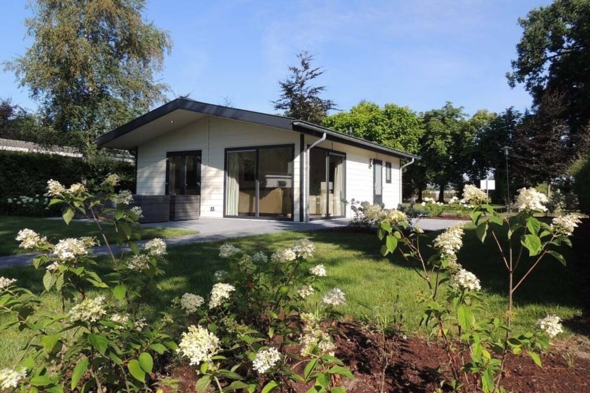 Uma casa moderna de dois andares com varanda coberta, um jardim da frente com plantas floridas e um céu azul límpido.