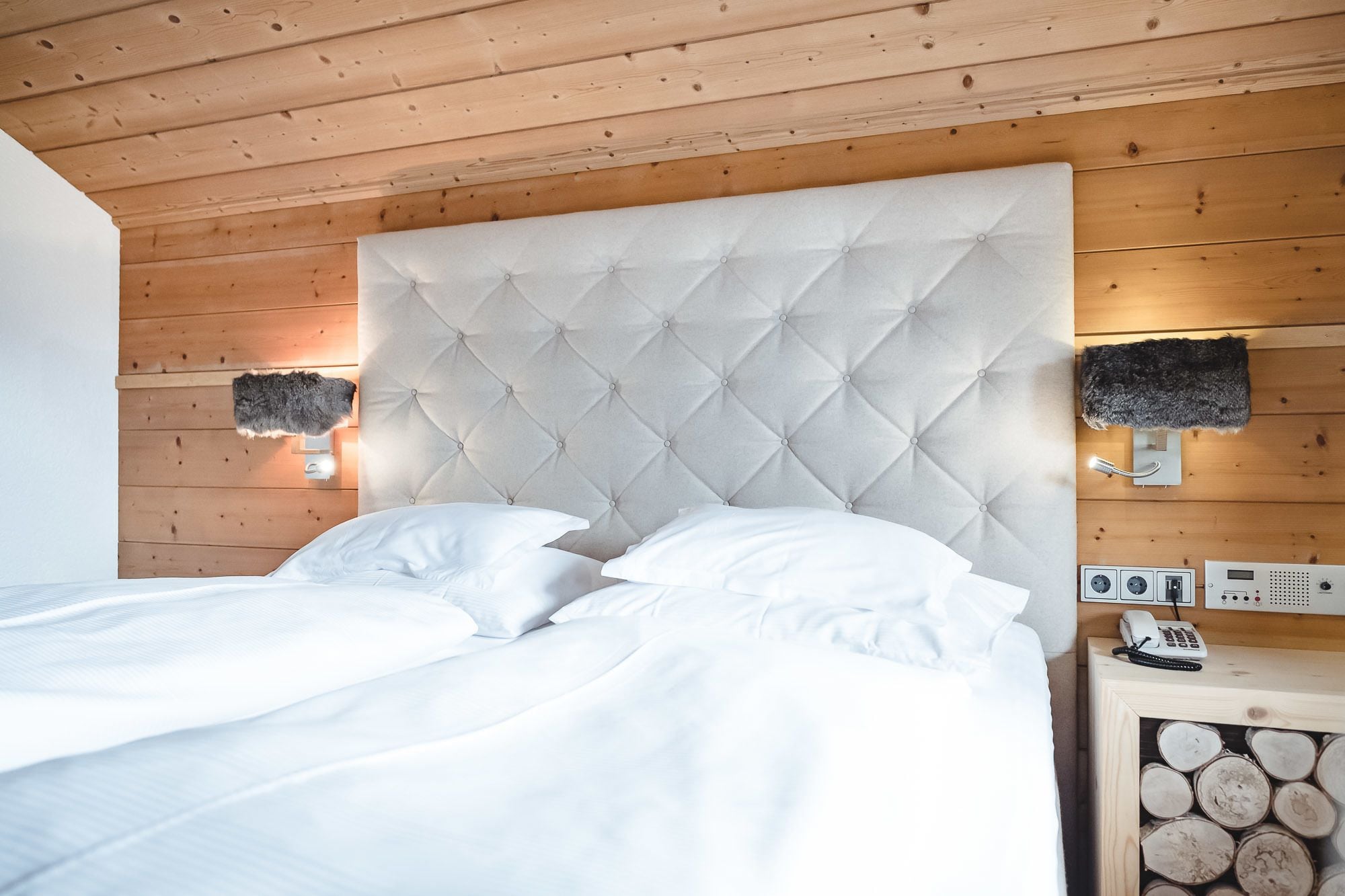 Quarto com cabeceira estofada, roupa de cama branca, mesa de cabeceira com telefone e luminária fixada na parede.