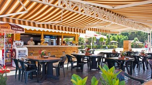 Um café ao ar livre com mesas e cadeiras de madeira, um balcão com cardápio e uma placa com a inscrição "CAFE ZERO".