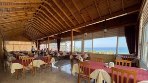 Uma ampla área de jantar com vigas de madeira no teto, mesas com toalhas vermelhas e brancas, e grandes janelas com vista para o oceano.