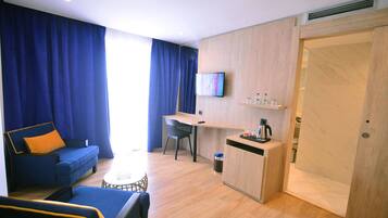 Quarto de hotel moderno com um sofá azul, uma mesinha de centro redonda, uma escrivaninha com cadeira, uma televisão e um minibar.