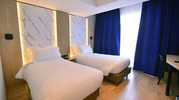 Quarto de hotel com duas camas, uma escrivaninha e uma janela grande com cortinas azuis.