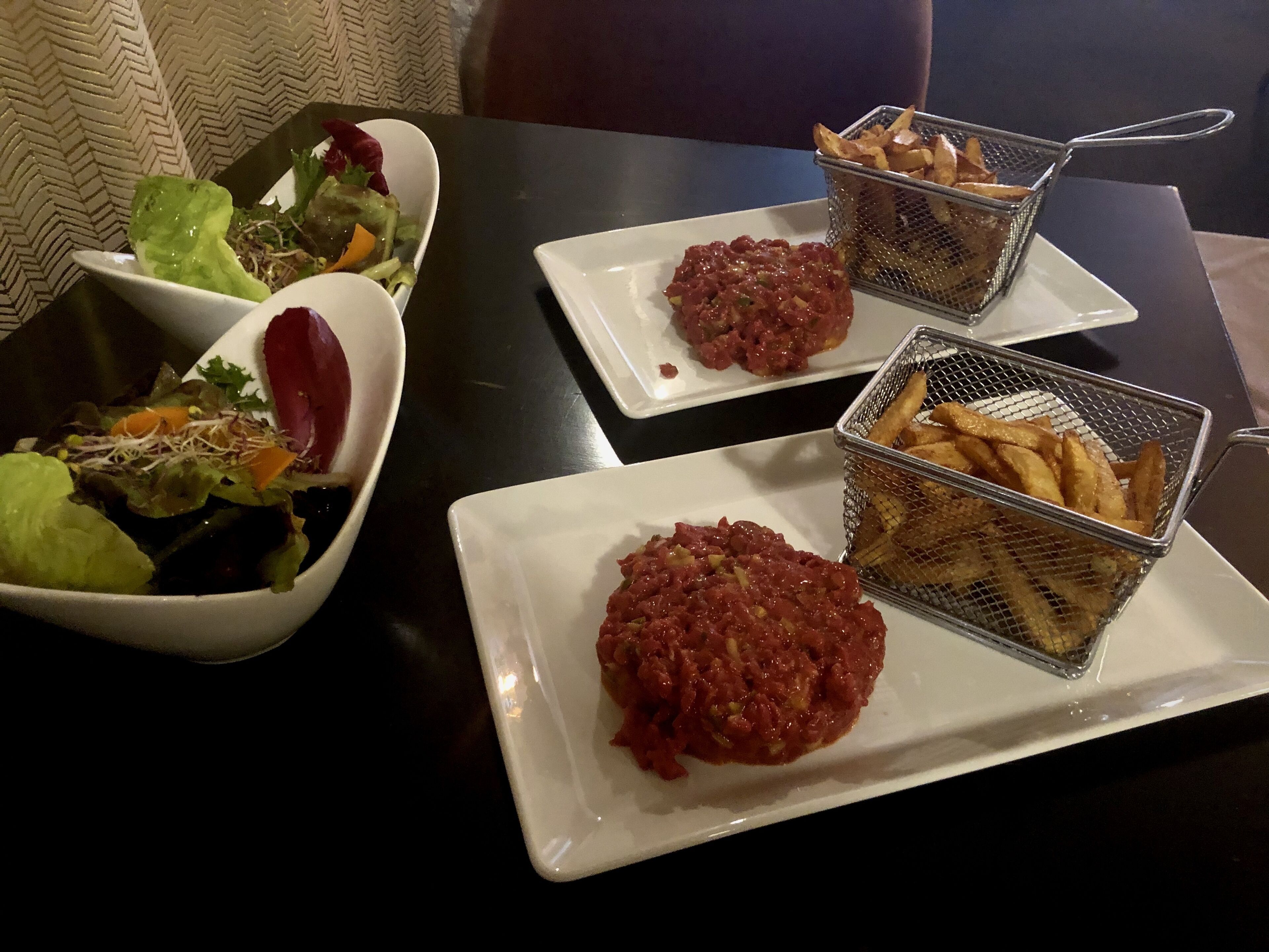 Uma mesa com três pratos brancos, cada um com um prato diferente: uma salada, um prato de carne vermelha e batatas fritas.