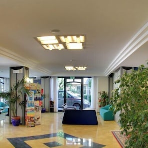 Um hall amplo com balcão de recepção, plantas em vasos e uma grande janela com vista para carros estacionados lá fora.