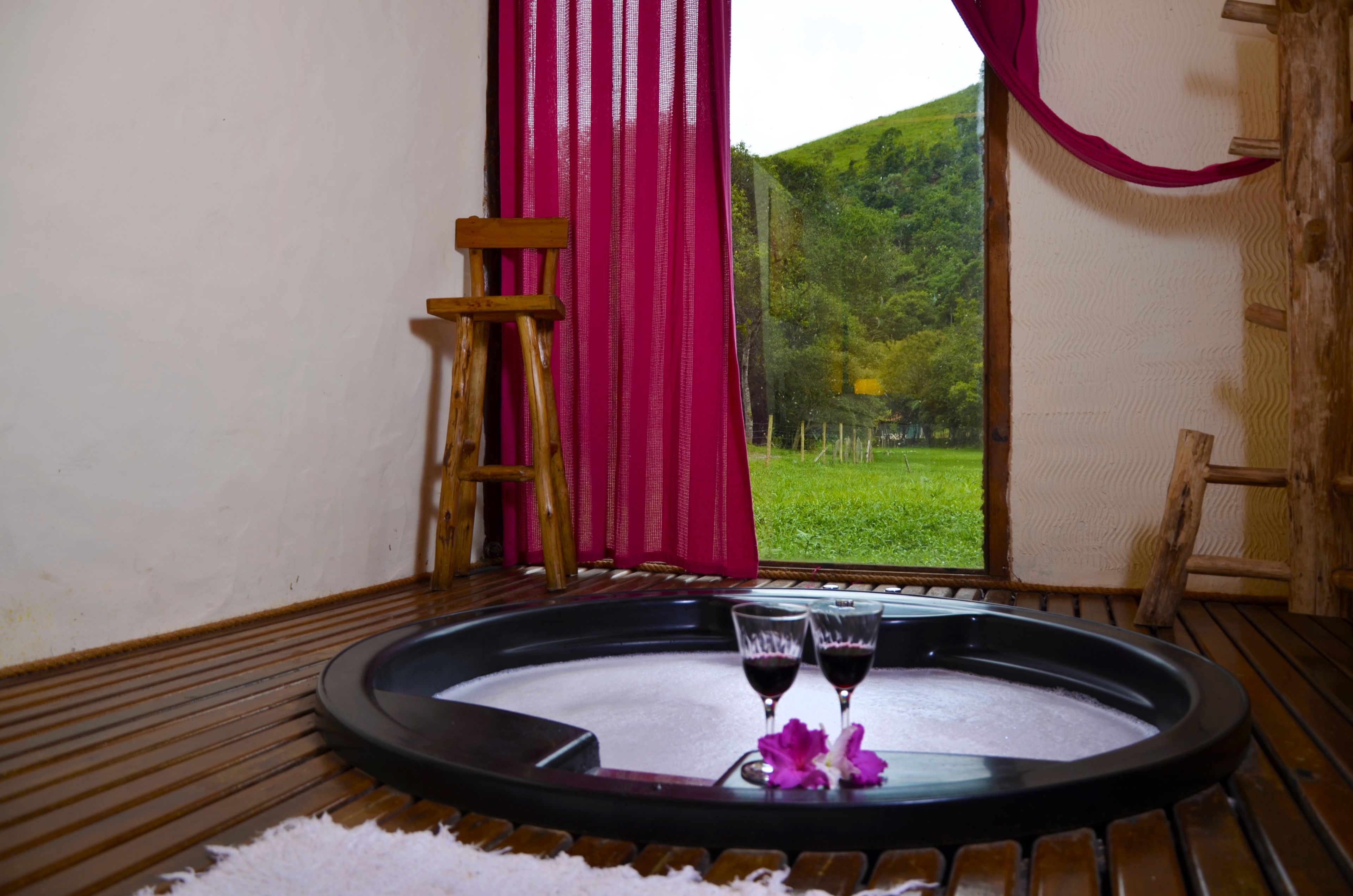 Quarto com piso de madeira, uma pequena cadeira de madeira, janela com cortinas cor-de-rosa e uma banheira com duas taças de vinho tinto e uma flor.