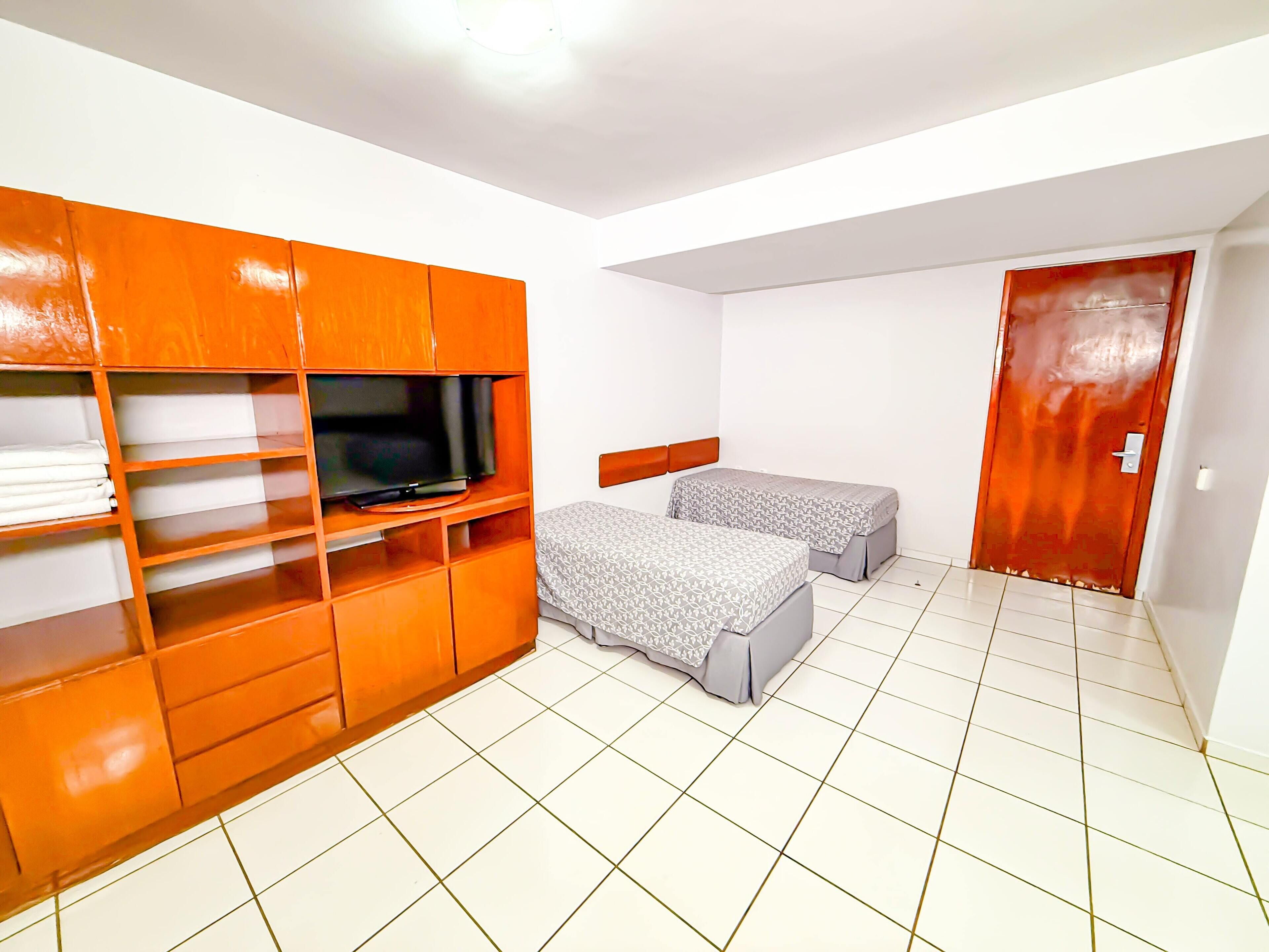 Quarto com televisão sobre um móvel de madeira, duas camas com roupa de cama estampada, uma porta de madeira e piso com azulejos brancos.