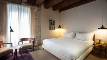 Quarto com uma cama grande, uma cadeira, uma mesa pequena, um abajur e um telefone.