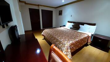 Quarto de hotel com cama, escrivaninha, cadeira, televisão e guarda-roupa.