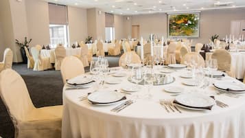 Salão de festas com mesas redondas preparadas para um evento formal, com toalhas brancas, talheres e copos.