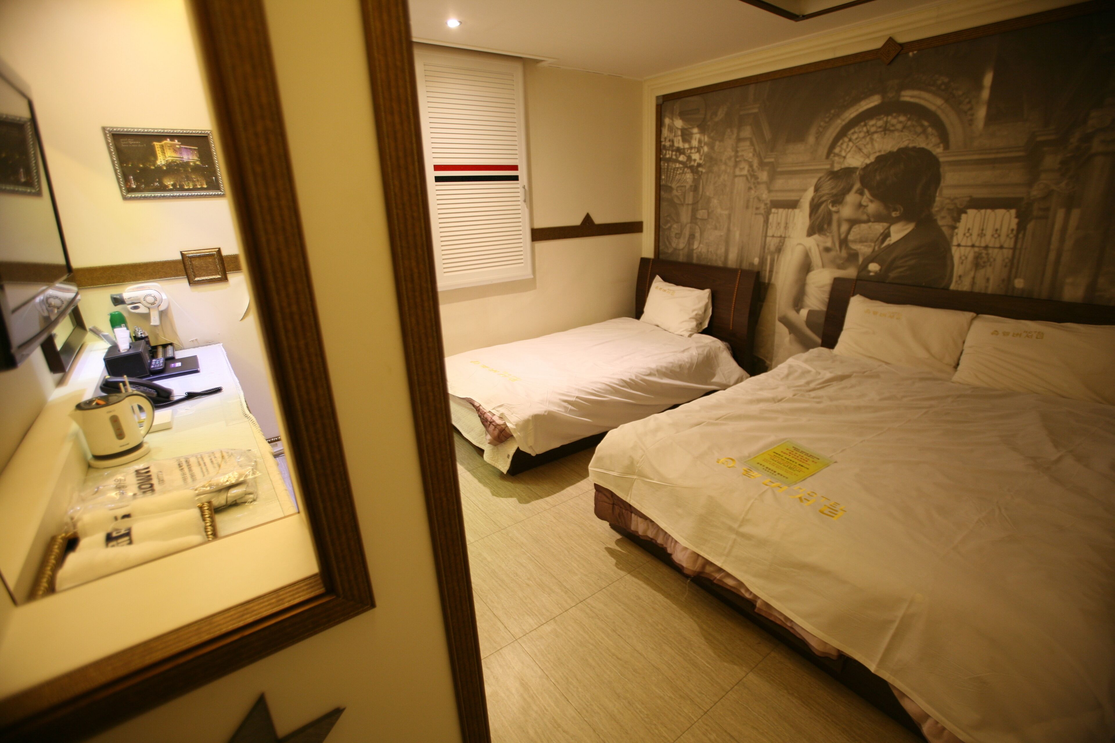 Quarto de hotel com duas camas, uma escrivaninha com telefone e máquina de café, janela com persianas e um quadro na parede.