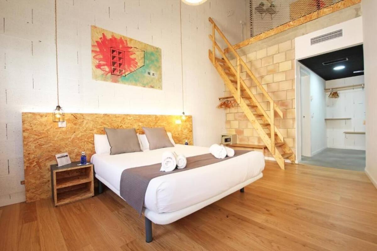 Quarto de hotel moderno com cabeceira de madeira, cama com roupa de cama branca, mesa de cabeceira de madeira, um quadro na parede e escada que leva a um mezanino.