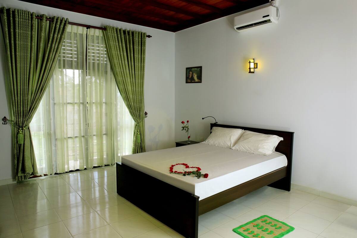 Quarto com cama, cortinas, ar-condicionado fixado na parede, mesa de cabeceira com um vaso e um tapete verde no chão.