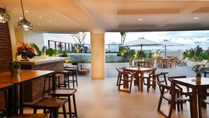 Uma área de refeições ao ar livre com mesas e cadeiras de madeira, um bar com balcão de tijolos e vista para o mar.