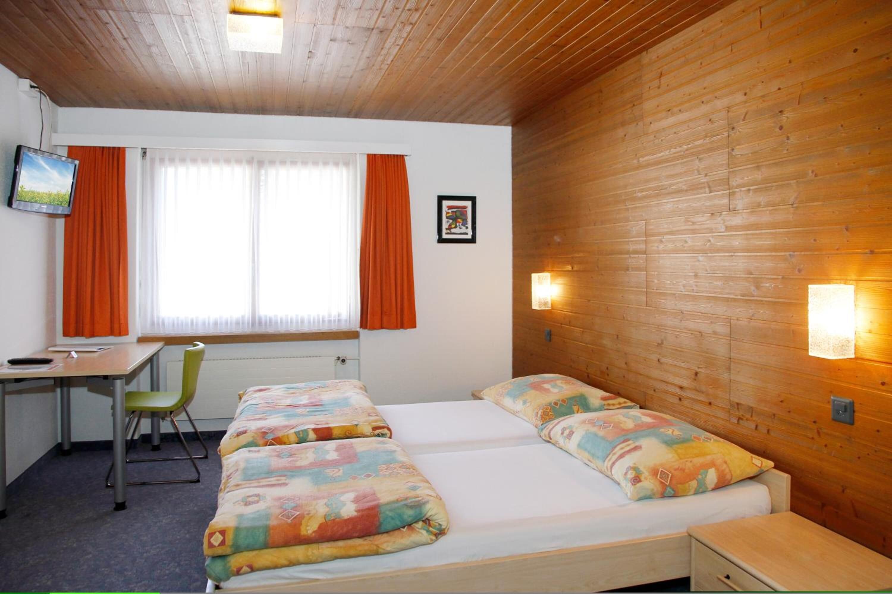 Quarto com duas camas, uma escrivaninha, uma cadeira e uma televisão na parede.