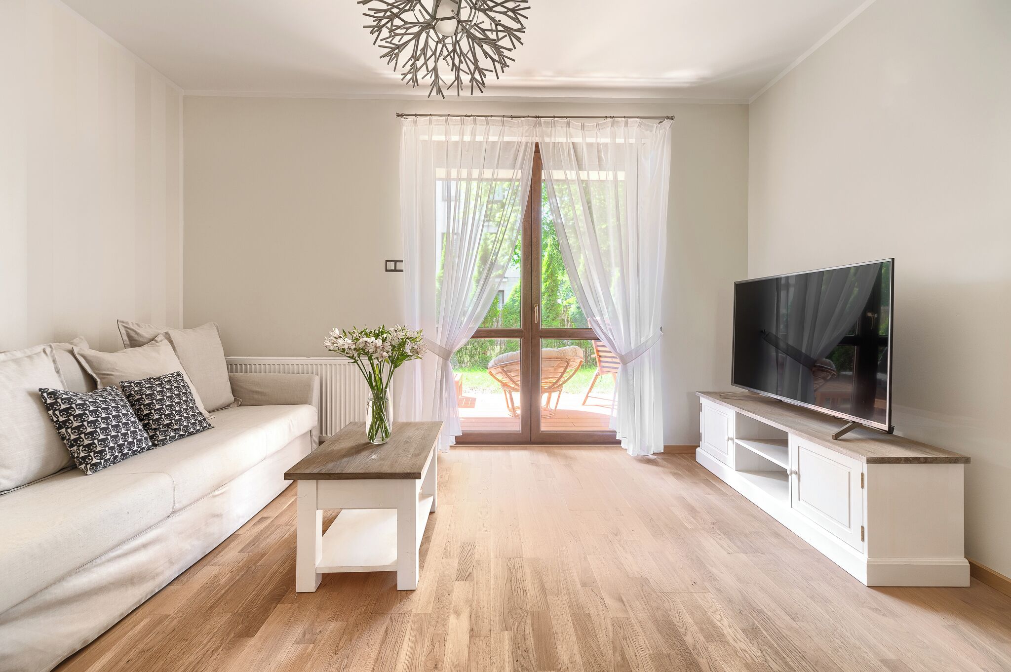 Uma sala de estar moderna com um sofá, uma mesa de centro, uma TV sobre um suporte e uma vista através de uma porta de vidro para uma área externa.