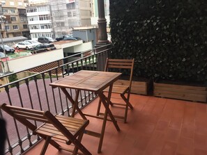 Uma varanda com mesa e cadeiras de madeira, grades de metal e vista para carros estacionados e edifícios ao fundo.