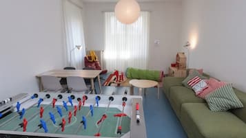 Um quarto com mesa de pebolim, sofá verde, uma pequena mesa com cadeiras e uma luminária.