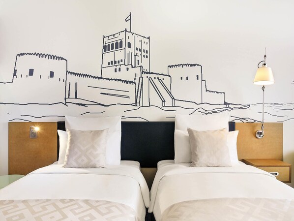 Quarto de hotel com duas camas, um mural na parede, uma mesinha de cabeceira e um abajur.