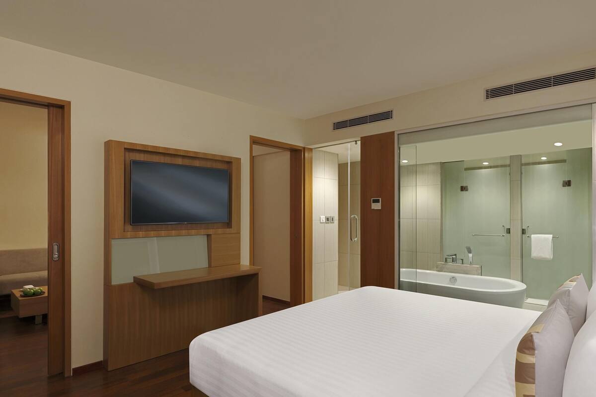 Quarto de hotel moderno com uma cama grande, televisão fixada na parede, escrivaninha de madeira, casa de banho com divisórias de vidro e banheira, e roupeiro com portas deslizantes.