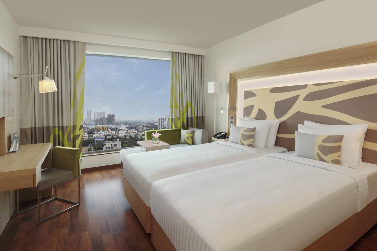 Quarto de hotel com uma cama grande, uma mesa e uma cadeira. Há uma janela com vista para a cidade.