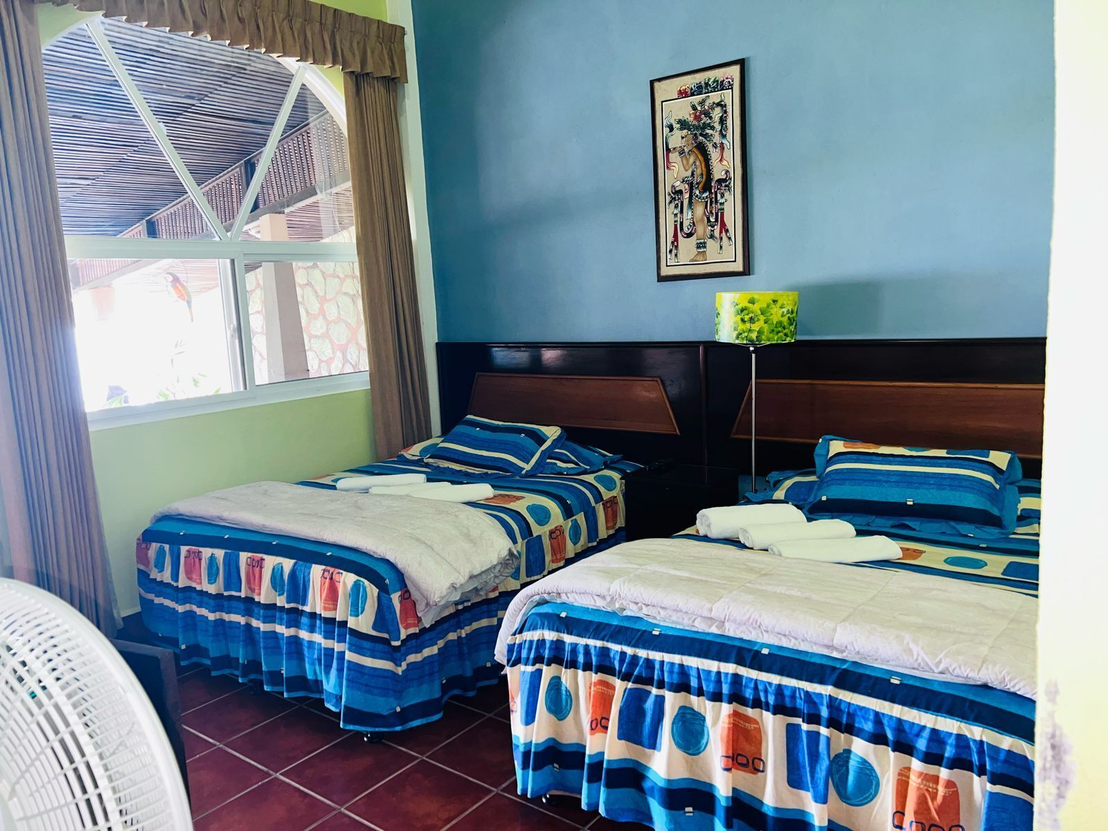 Quarto de hotel com duas camas, uma janela com cortinas, um ventilador e um quadro na parede.